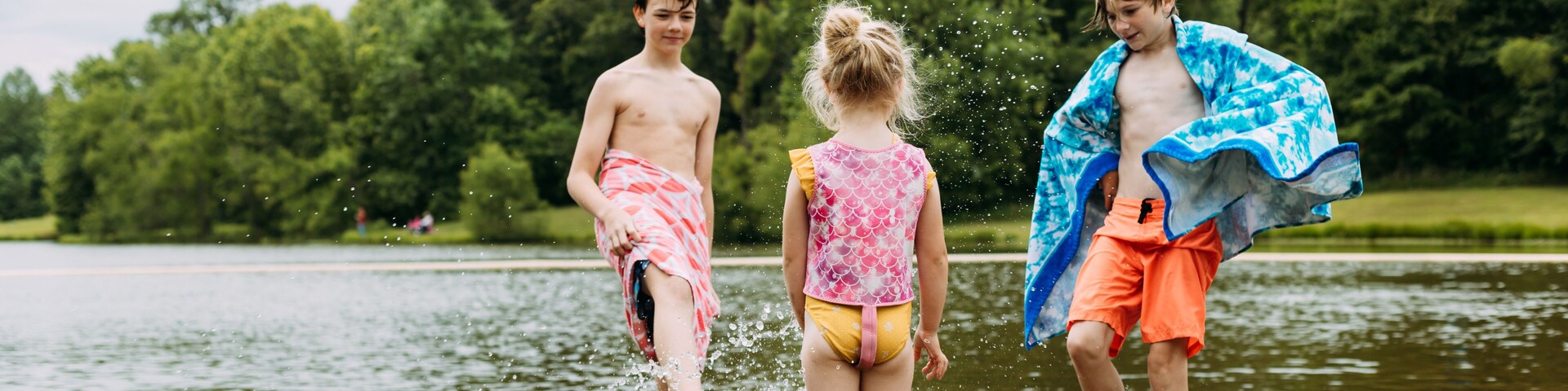 Três crianças brincando à beira de um lago, com árvores ao fundo.
