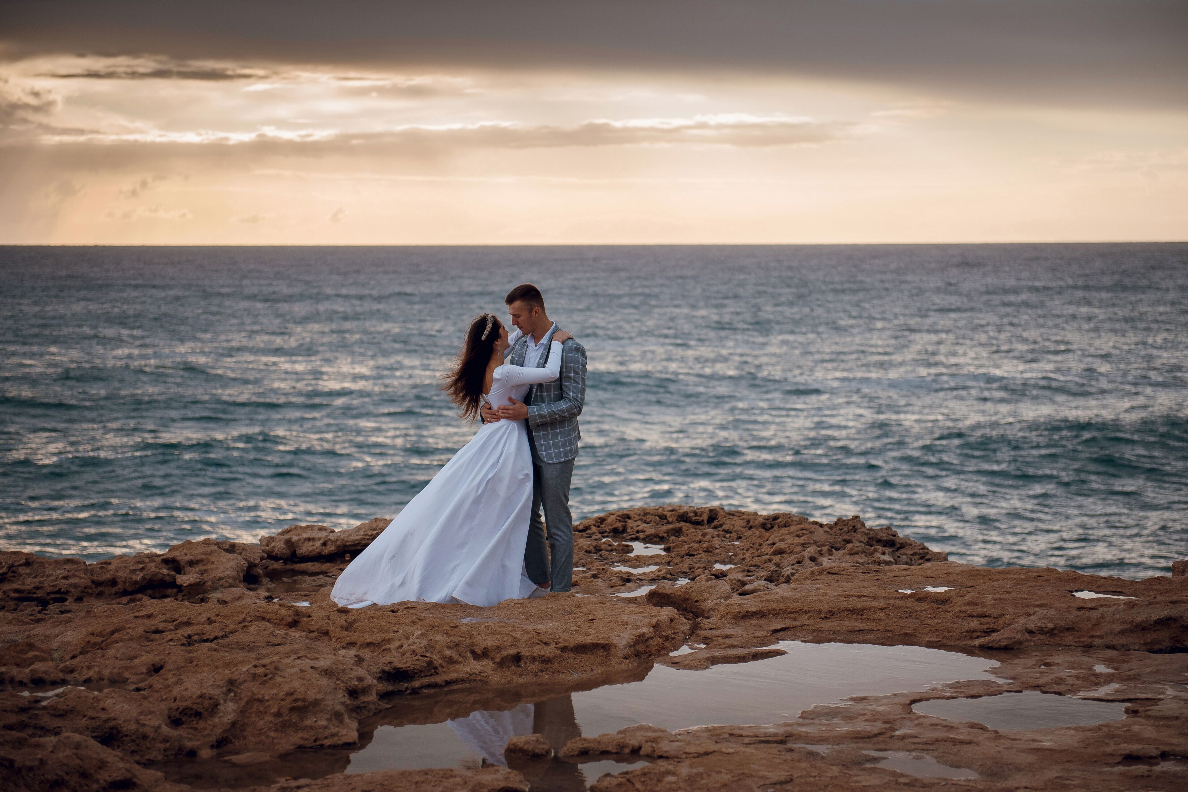 Um casal se abraça em uma praia rochosa, com o oceano ao fundo.