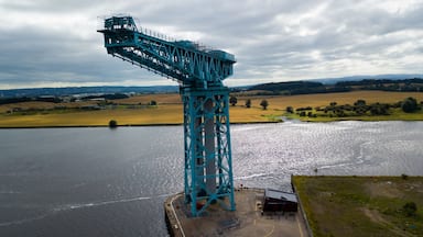 Uma grande estrutura de torre guindaste azul sobre um rio, com campos e colinas ao fundo.