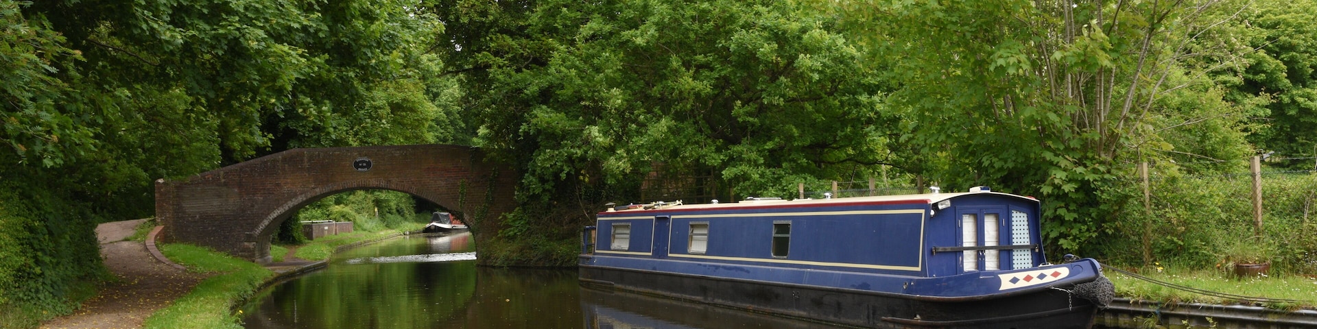 Um canal estreito com um barco azul atracado à direita, uma ponte de tijolos ao fundo e árvores verdes densas ao longo do canal.