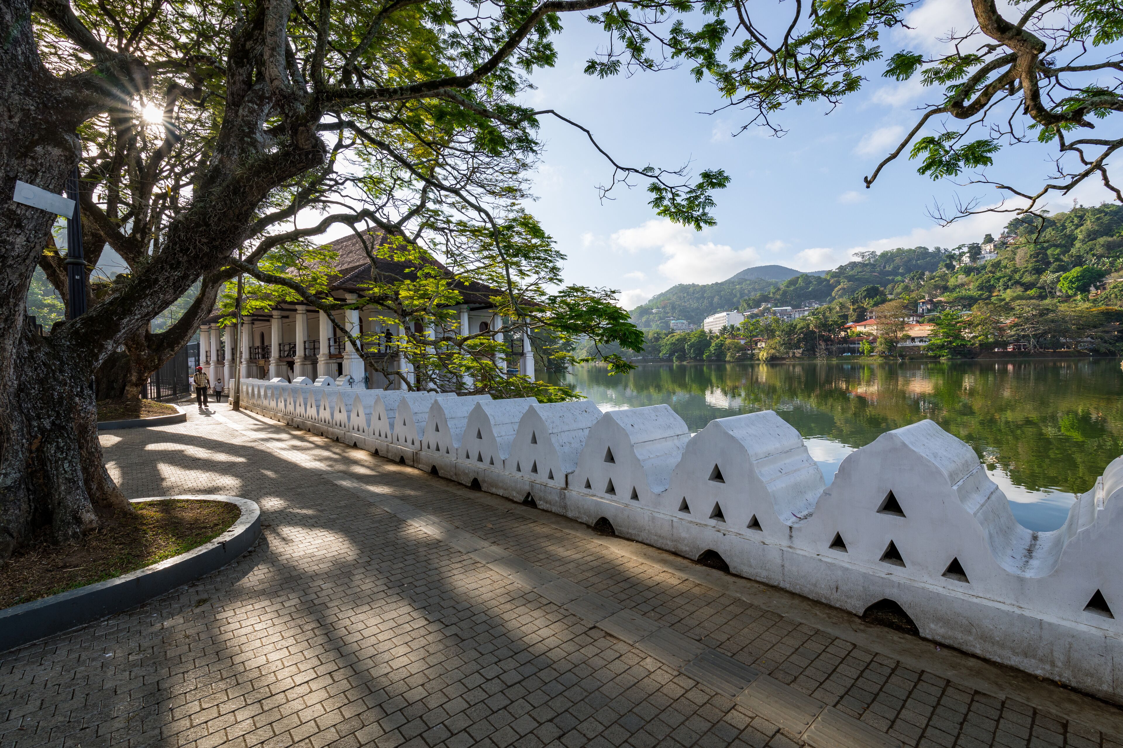 Uma passarela pavimentada ao lado de um lago calmo, com um corrimão decorativo branco e árvores oferecendo sombra.