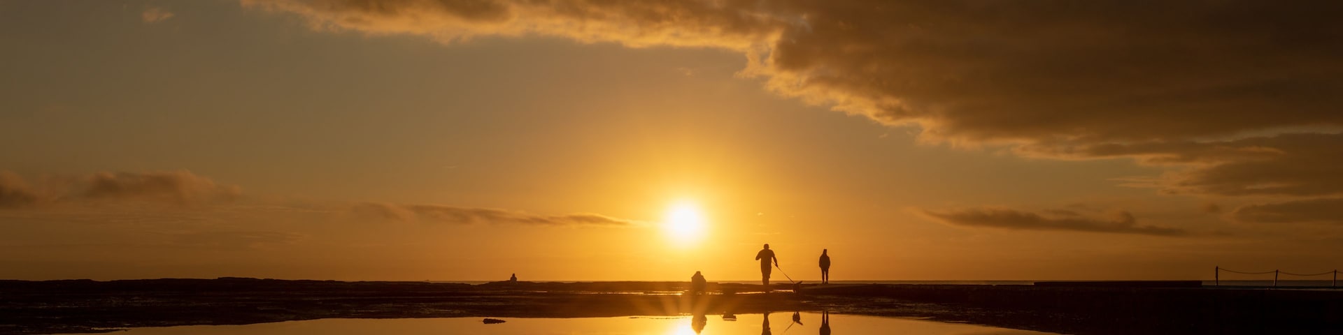 Pôr do sol refletido na água, com silhuetas de pessoas e uma linha de costa distante.