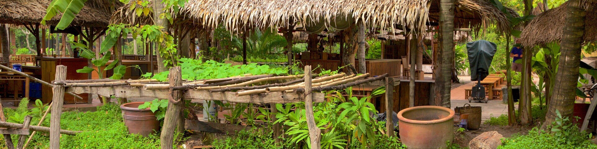 Uma construção rústica com telhado de palha, rodeada por vegetação exuberante e um corpo d'água calmo.