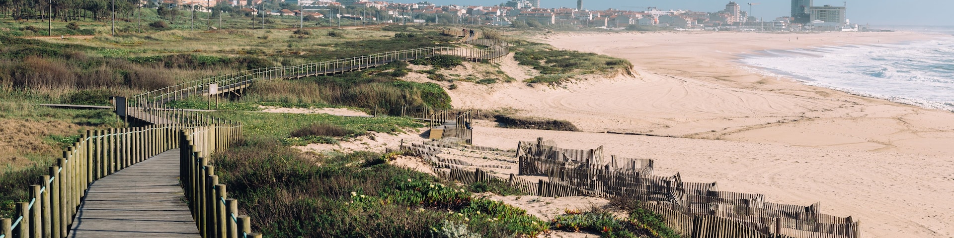 Um calçadão de madeira ao longo de uma praia arenosa, com dunas e vegetação.