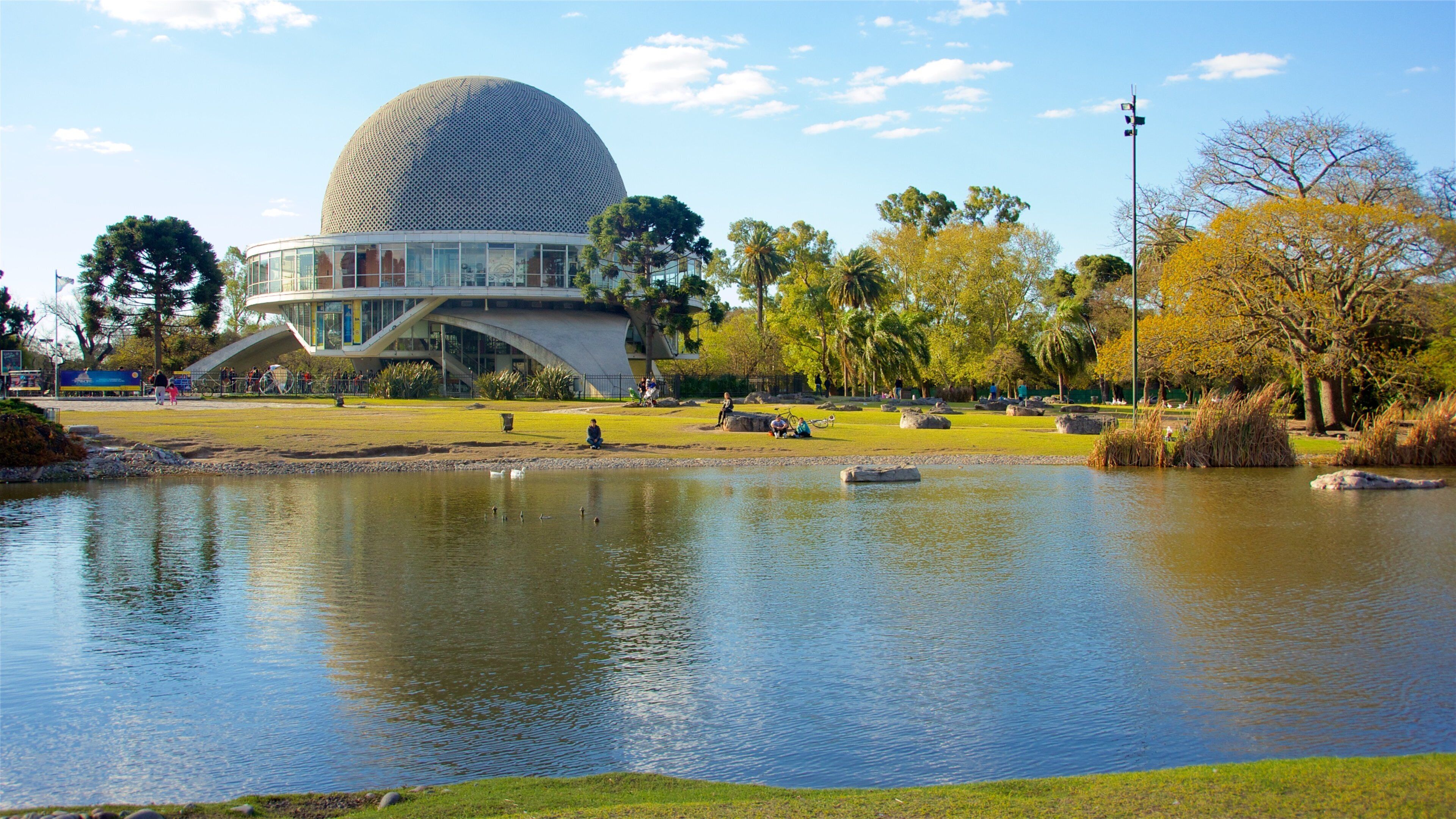 Um edifício moderno com uma cúpula esférica, um lago com patos e um parque com pessoas e árvores.