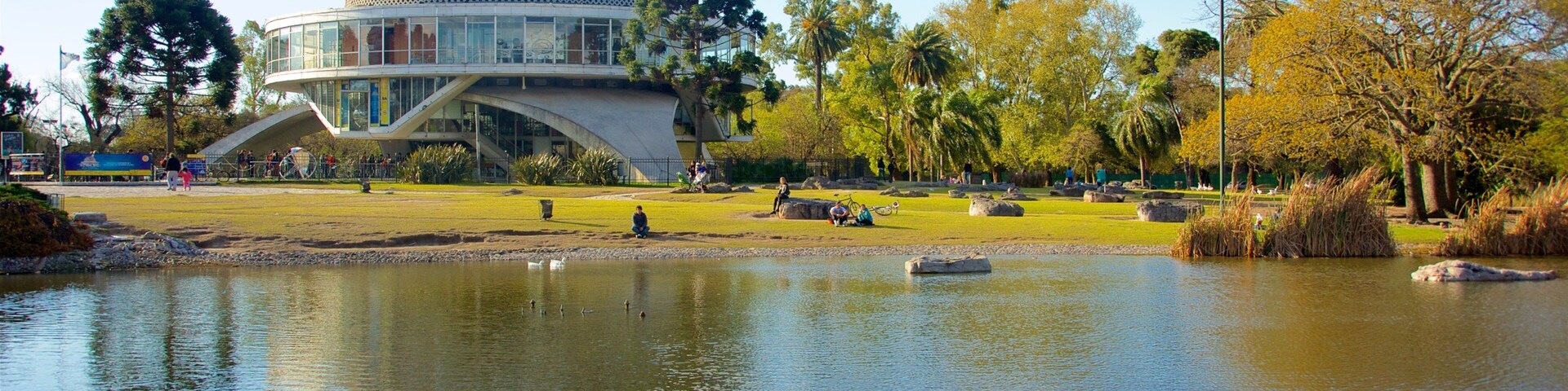 Um edifício moderno com uma cúpula esférica, um lago com patos e um parque com pessoas e árvores.