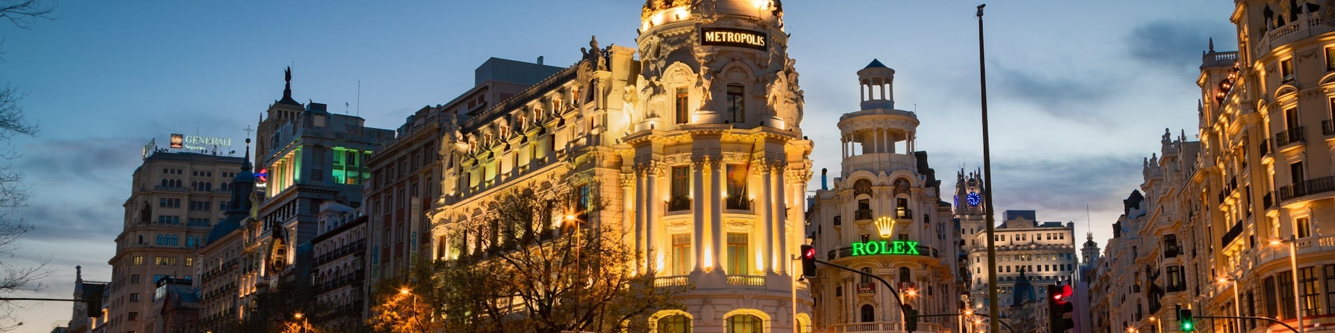 Um edifício histórico com arquitetura ornamentada e uma cúpula, rodeado por edifícios modernos e uma rua movimentada com rastros de luz deixados por veículos em movimento.