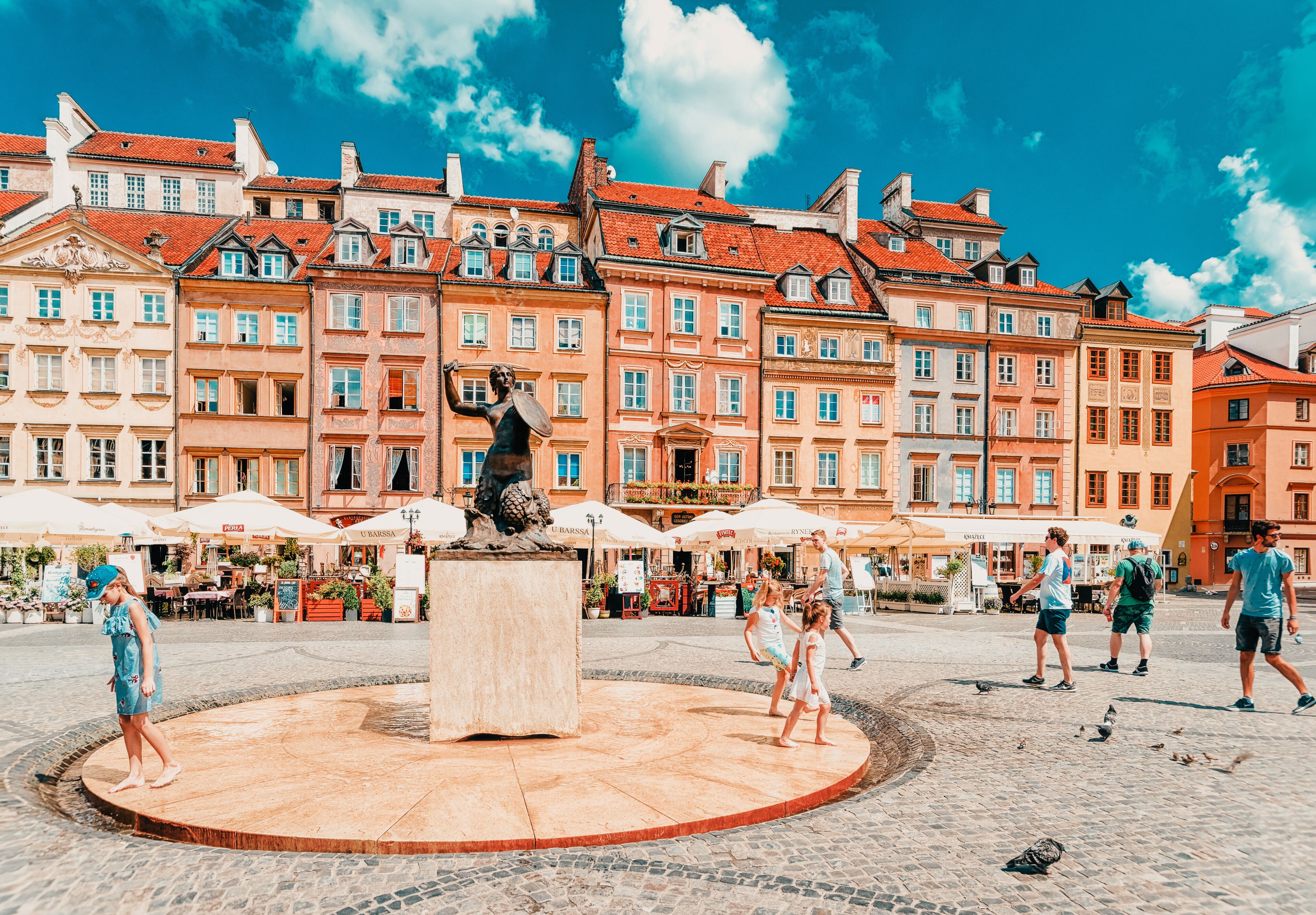 Uma praça histórica com edifícios ornamentados, uma fonte central e áreas de estar ao ar livre.