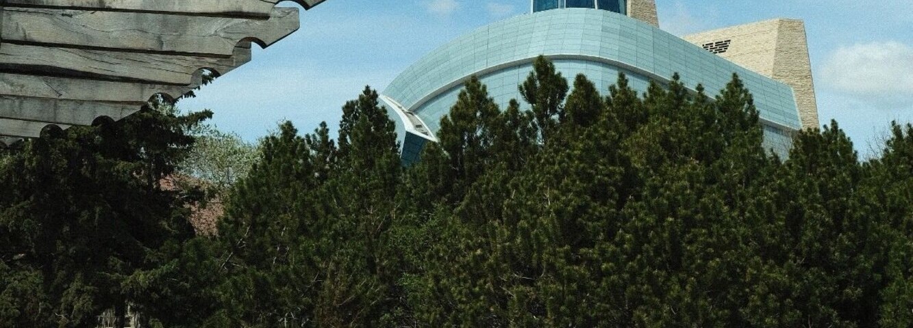 Um edifício moderno com uma torre, rodeado por árvores e um caminho de concreto.