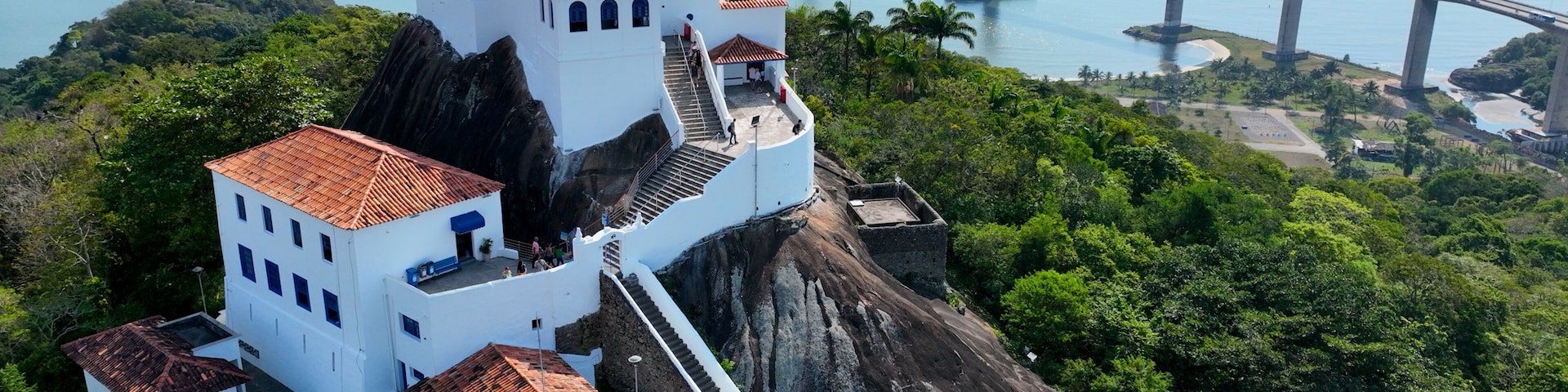Um castelo branco com telhado vermelho empoleirado em um penhasco com vista para uma cidade costeira e uma ponte.
