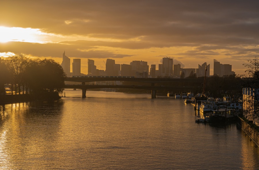 Um pôr do sol sobre o horizonte urbano, com uma ponte e barcos na água.