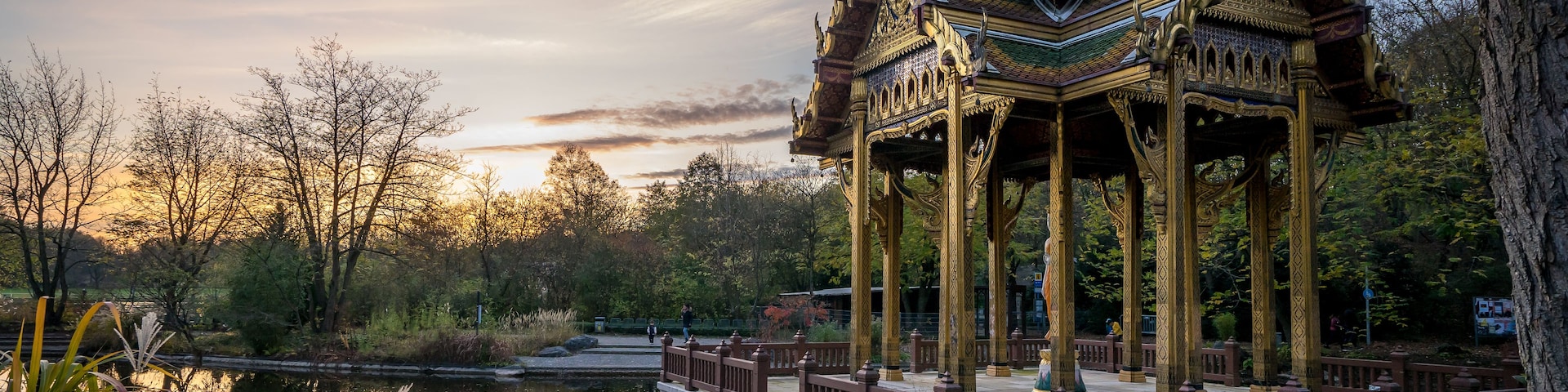 Um pavilhão tradicional com telhado dourado ricamente decorado e colunas, cercado por um lago tranquilo e árvores.