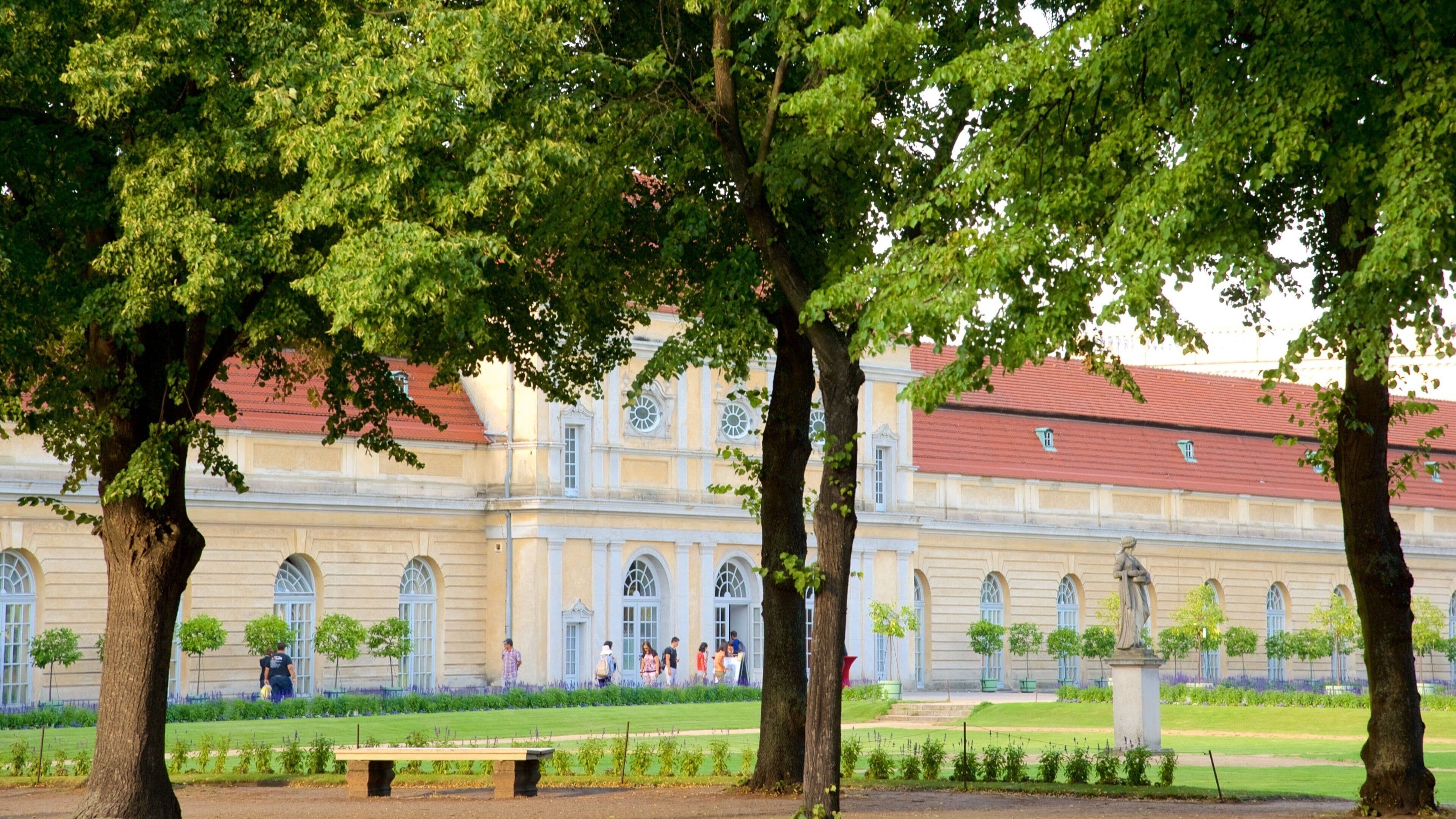 Um edifício histórico com janelas em arco e telhado vermelho, rodeado por árvores e um jardim bem cuidado.