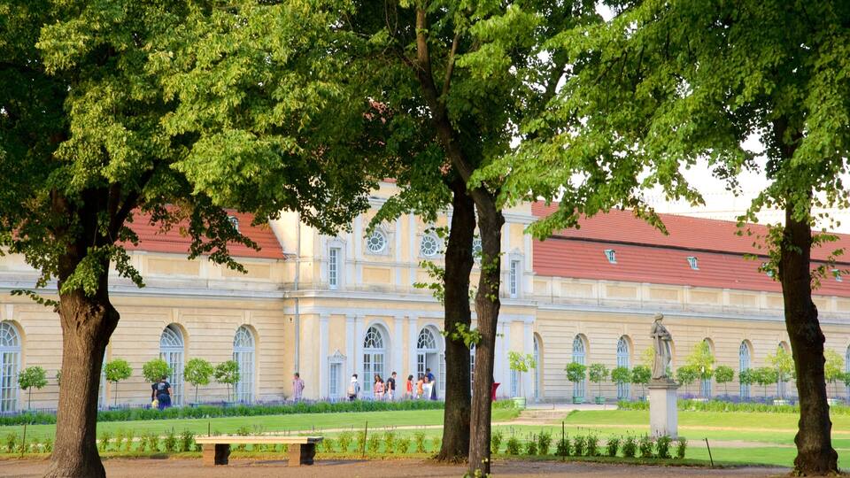 Um edifício histórico com janelas em arco e telhado vermelho, rodeado por árvores e um jardim bem cuidado.