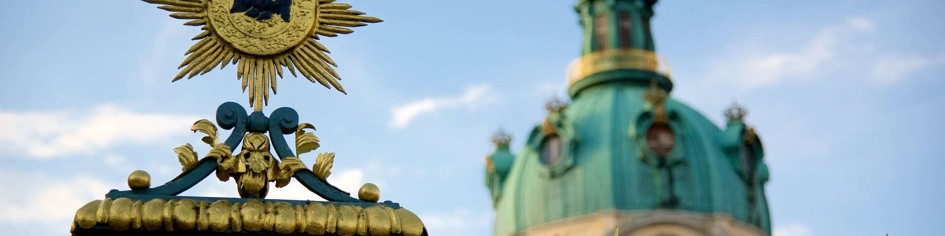 Emblema dourado do sol e portão ornamentado em primeiro plano, com um edifício de cúpula verde ao fundo.