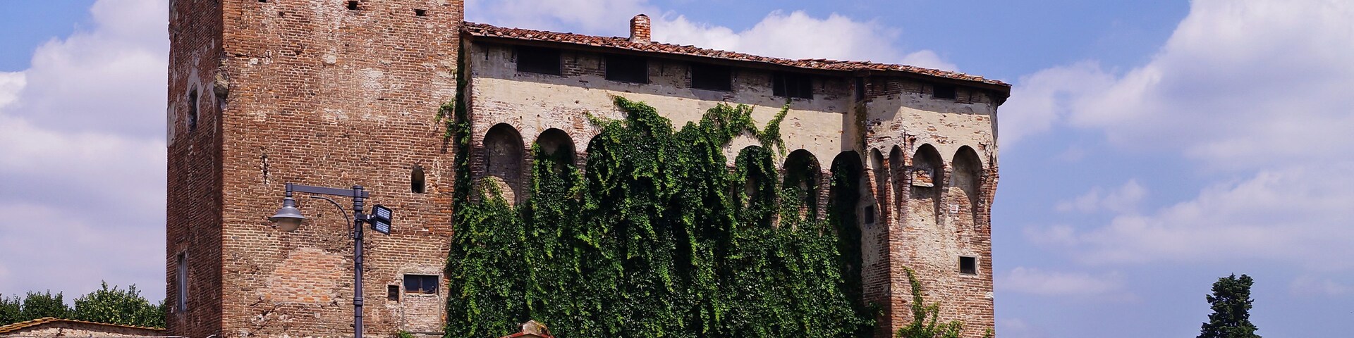 Um edifício histórico com uma torre alta e janelas em arco, cercado por vegetação e um céu claro.