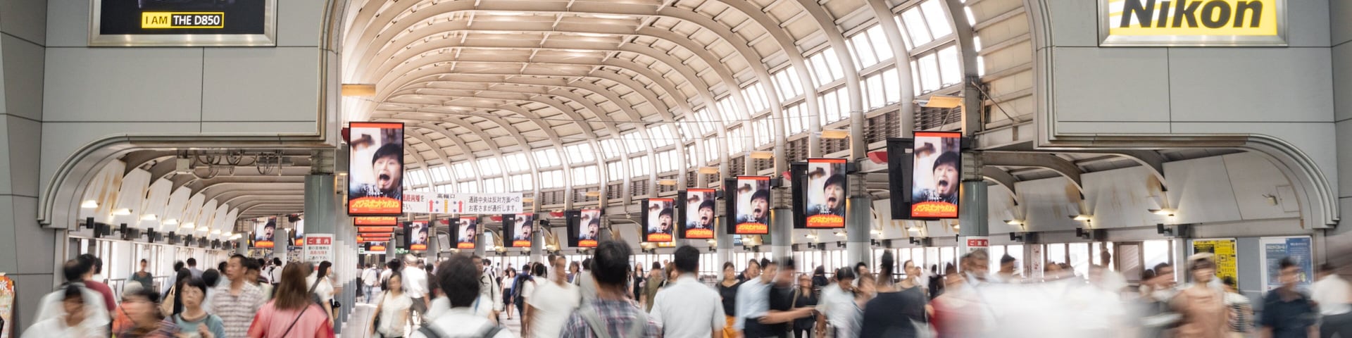 Uma estação de trem interna movimentada, com tetos arredondados e várias telas digitais exibindo anúncios.