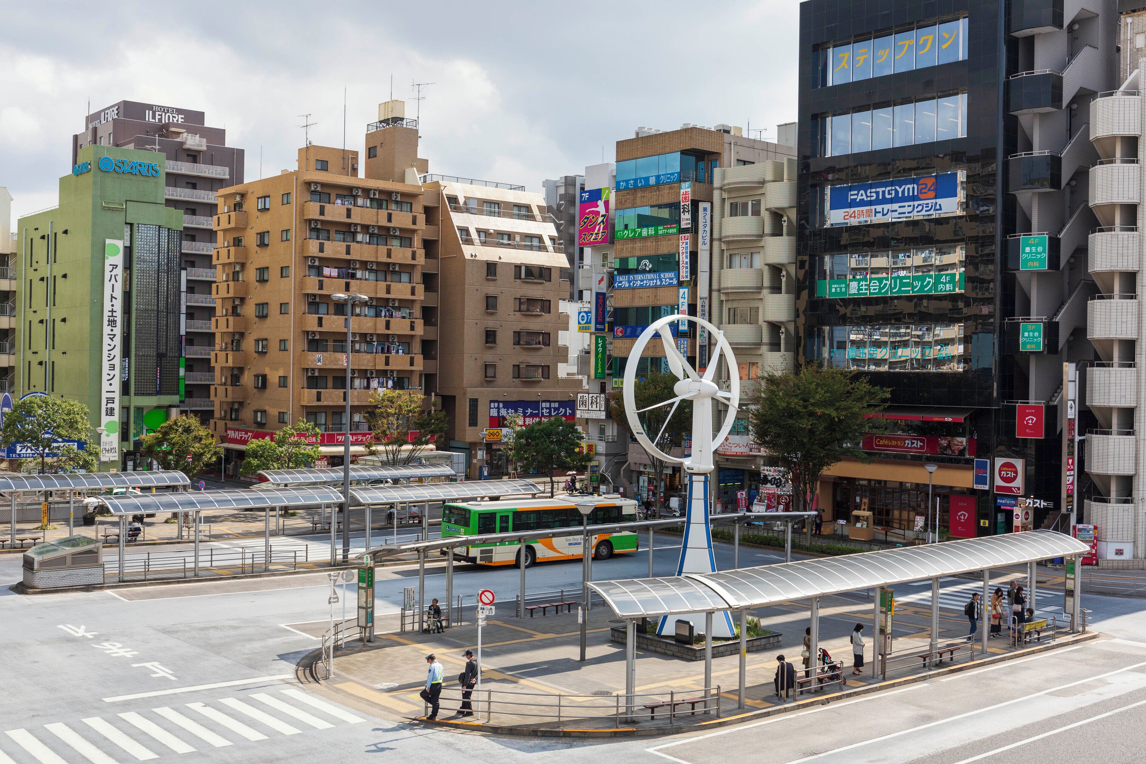 Um movimentado cruzamento urbano com uma escultura circular branca marcante, um ônibus verde e diversos edifícios comerciais.