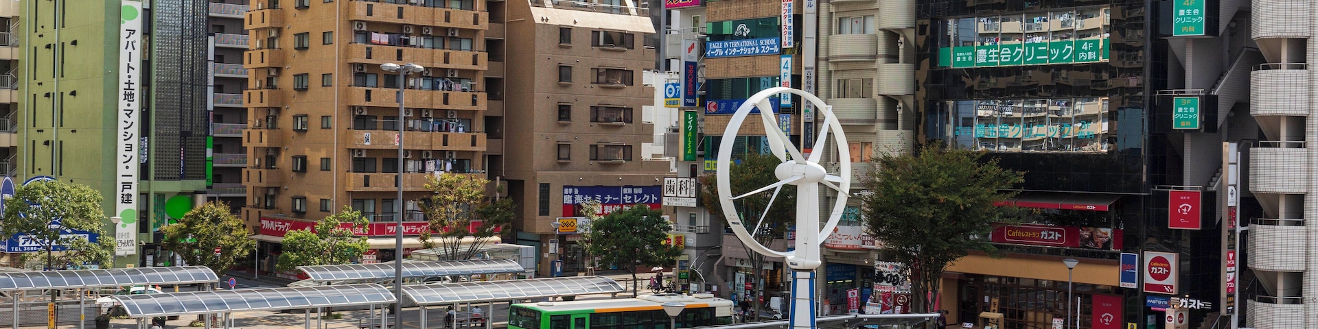 Um movimentado cruzamento urbano com uma escultura circular branca marcante, um ônibus verde e diversos edifícios comerciais.