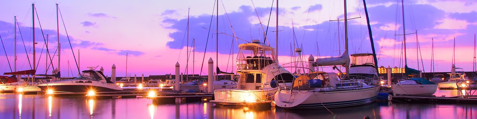 Um porto de iates ao entardecer, com vários veleiros atracados refletindo as cores do céu.