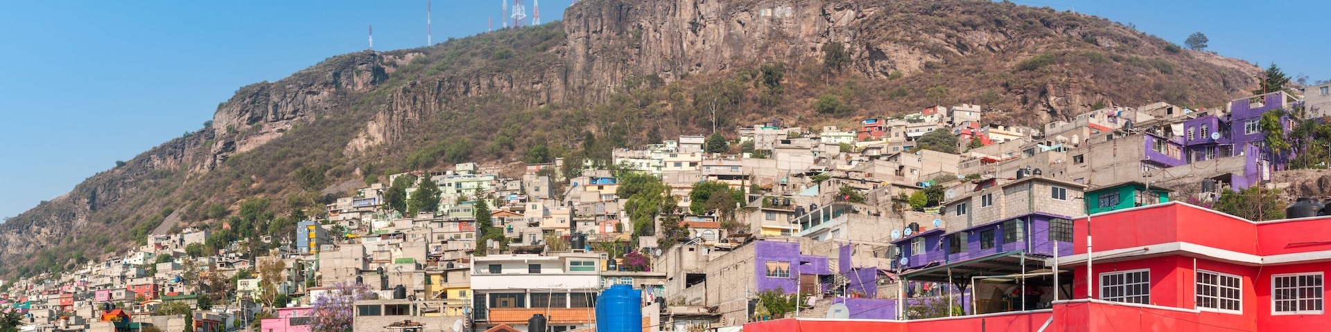Uma cidade pitoresca e vibrante, construída em uma encosta íngreme, com edifícios coloridos, uma colina rochosa imponente e algumas torres de comunicação ao fundo.