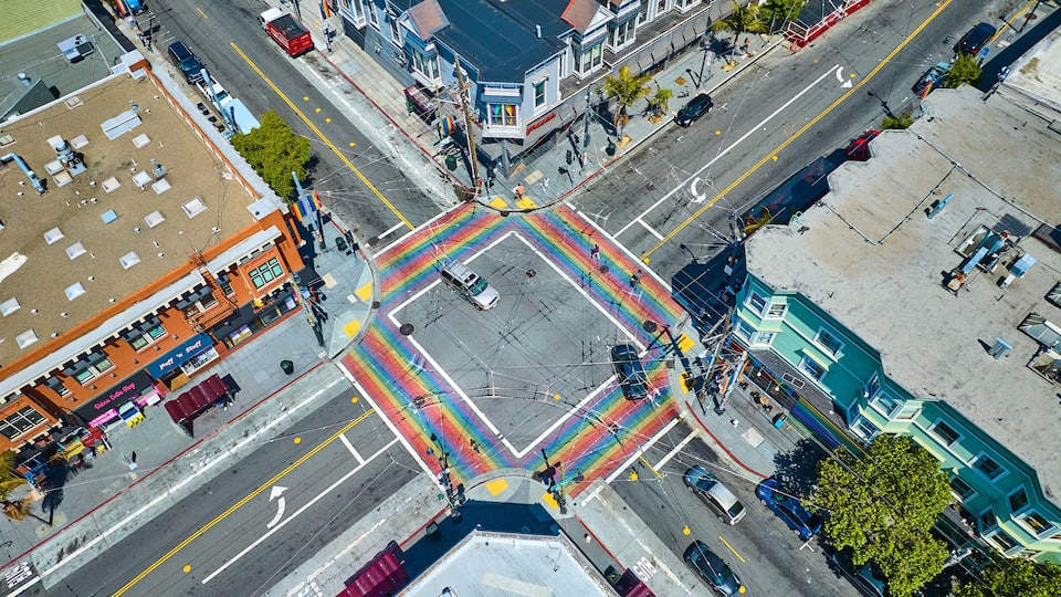 Vista aérea de um cruzamento urbano com uma faixa de pedestres colorida em arco-íris e edifícios ao redor.