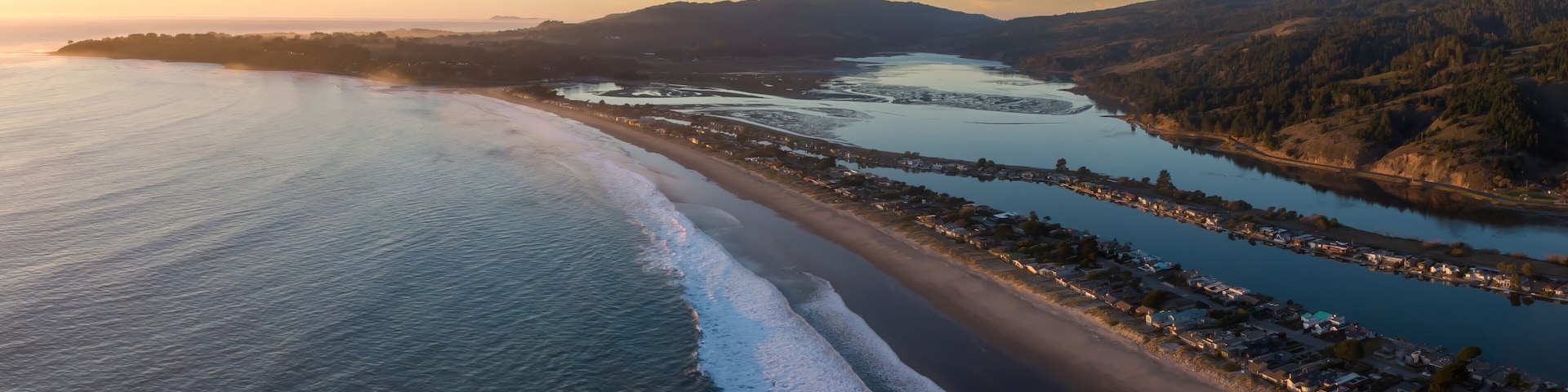 Paisagem costeira com uma praia, um rio e colinas ao fundo.