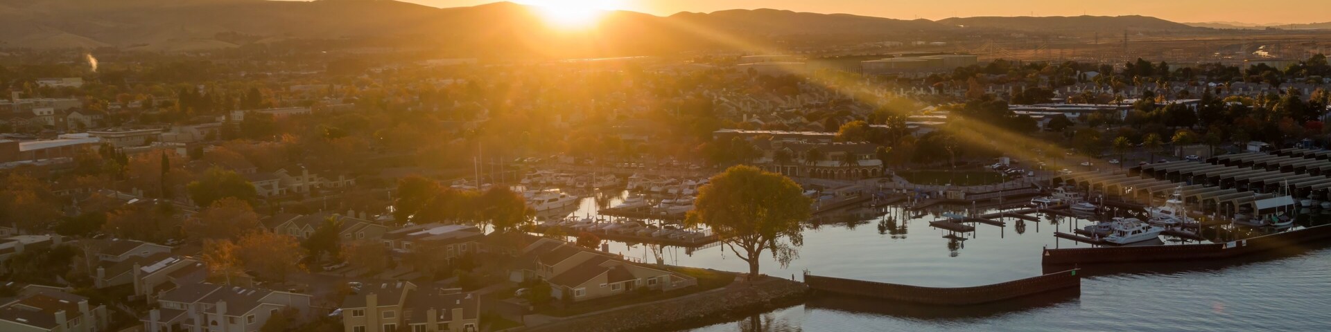 Pôr do sol sobre uma área residencial com um porto de embarcações e água calma refletindo o céu.