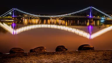 Uma ponte iluminada à noite refletindo em águas calmas, com pedras em primeiro plano.