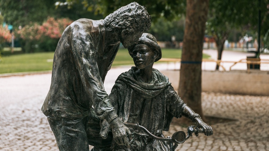 Uma estátua retrata uma pessoa ajudando outra em uma bicicleta, em um parque.