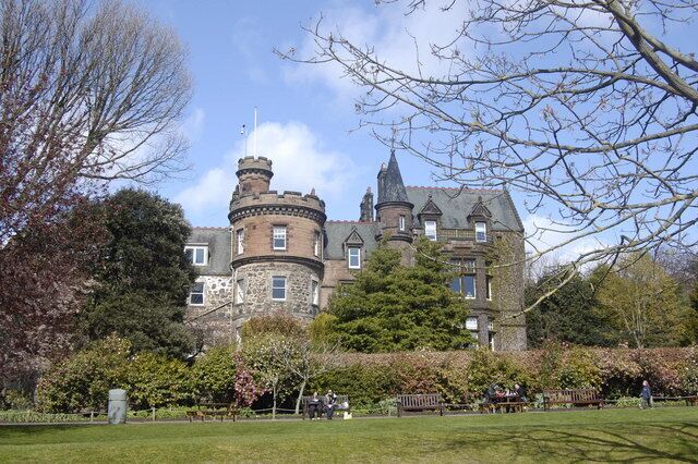 Um edifício grande e histórico com uma torre e várias chaminés, rodeado por um jardim bem cuidado e árvores.