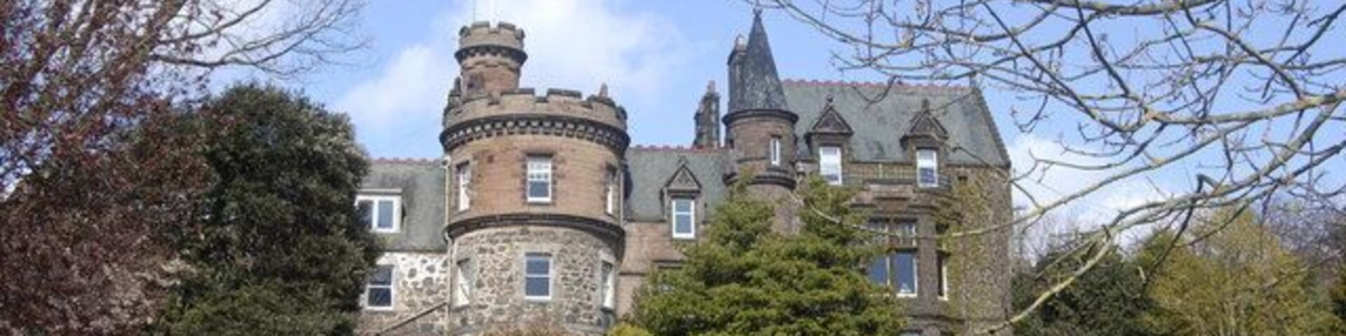 Um edifício grande e histórico com uma torre e várias chaminés, rodeado por um jardim bem cuidado e árvores.