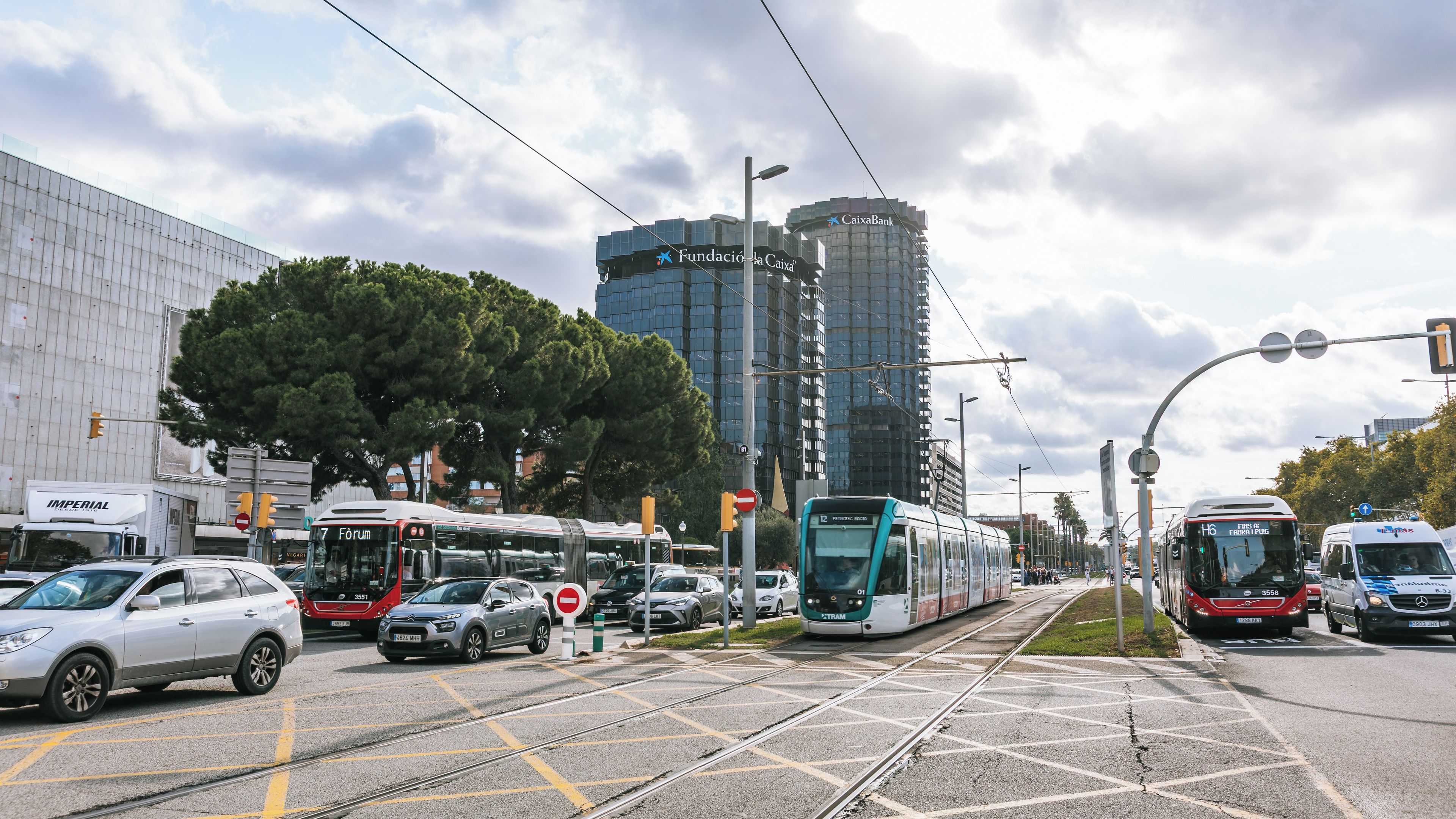 Um paragem de elétrico com um elétrico, carros e um edifício com a inscrição 'Caixabank'.