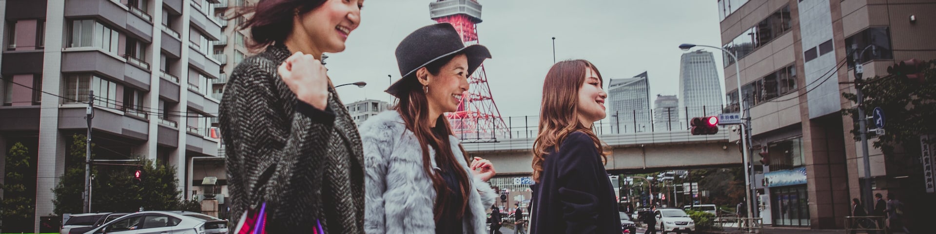 Três mulheres atravessam uma rua carregando sacolas de compras, com uma torre vermelha ao fundo e edifícios modernos.