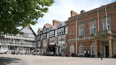 Uma praça histórica com edifícios em estilo Tudor, uma placa indicando Margem do Rio e pessoas caminhando.