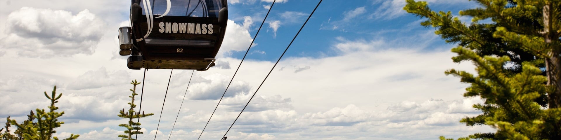 Um teleférico com a inscrição "SNOWMASS" suspenso entre árvores, com uma paisagem montanhosa ao fundo.