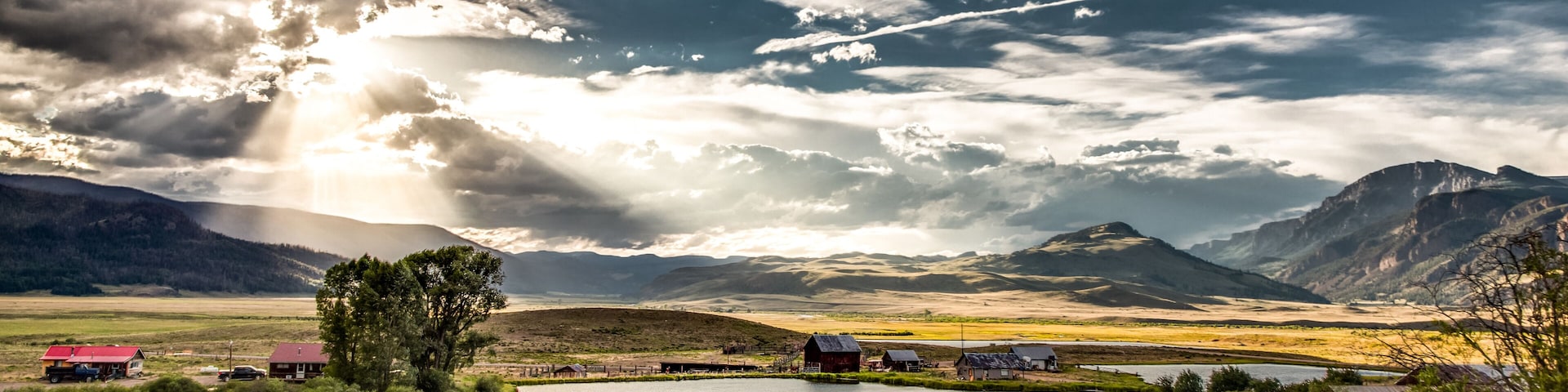 Paisagem rural com um lago, cabanas com telhados vermelhos e montanhas sob um céu nublado.
