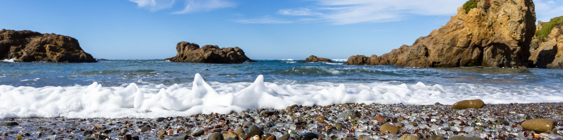 Uma praia de pedras com ondas, céu claro e formações rochosas.