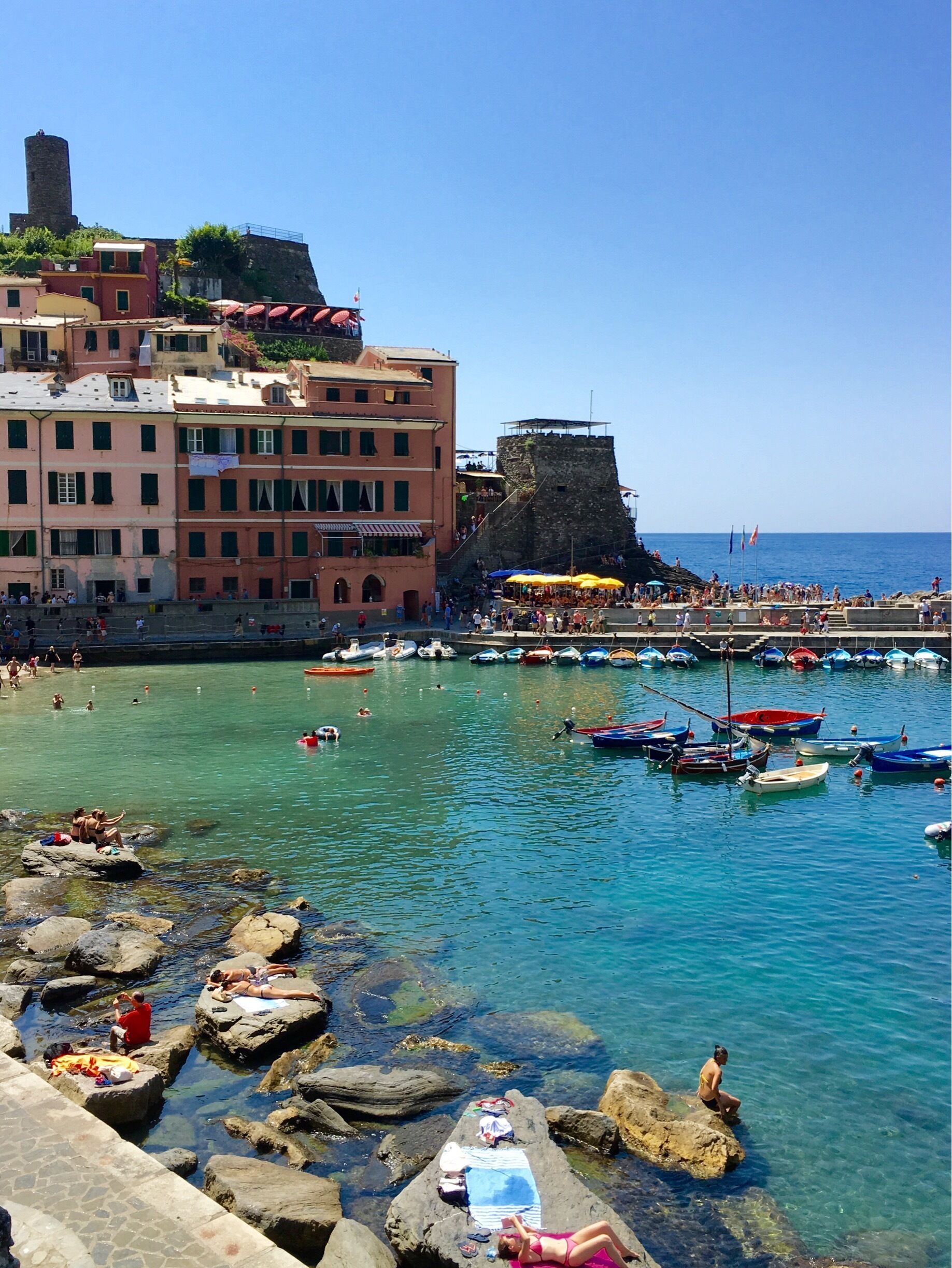 Uma vila costeira com edifícios coloridos, uma torre histórica e uma marina com barcos.