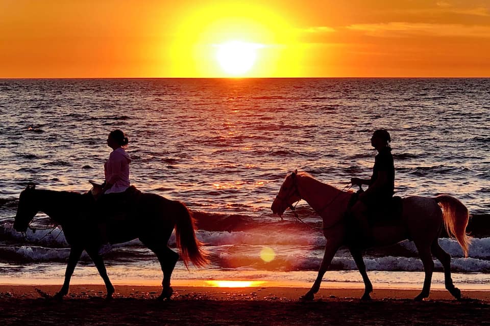 Duas pessoas cavalgam a cavalo ao longo de uma praia ao pôr do sol.
