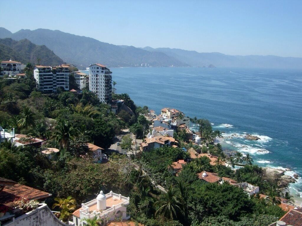 Uma vila costeira com edifícios, palmeiras e vista para o oceano e montanhas.