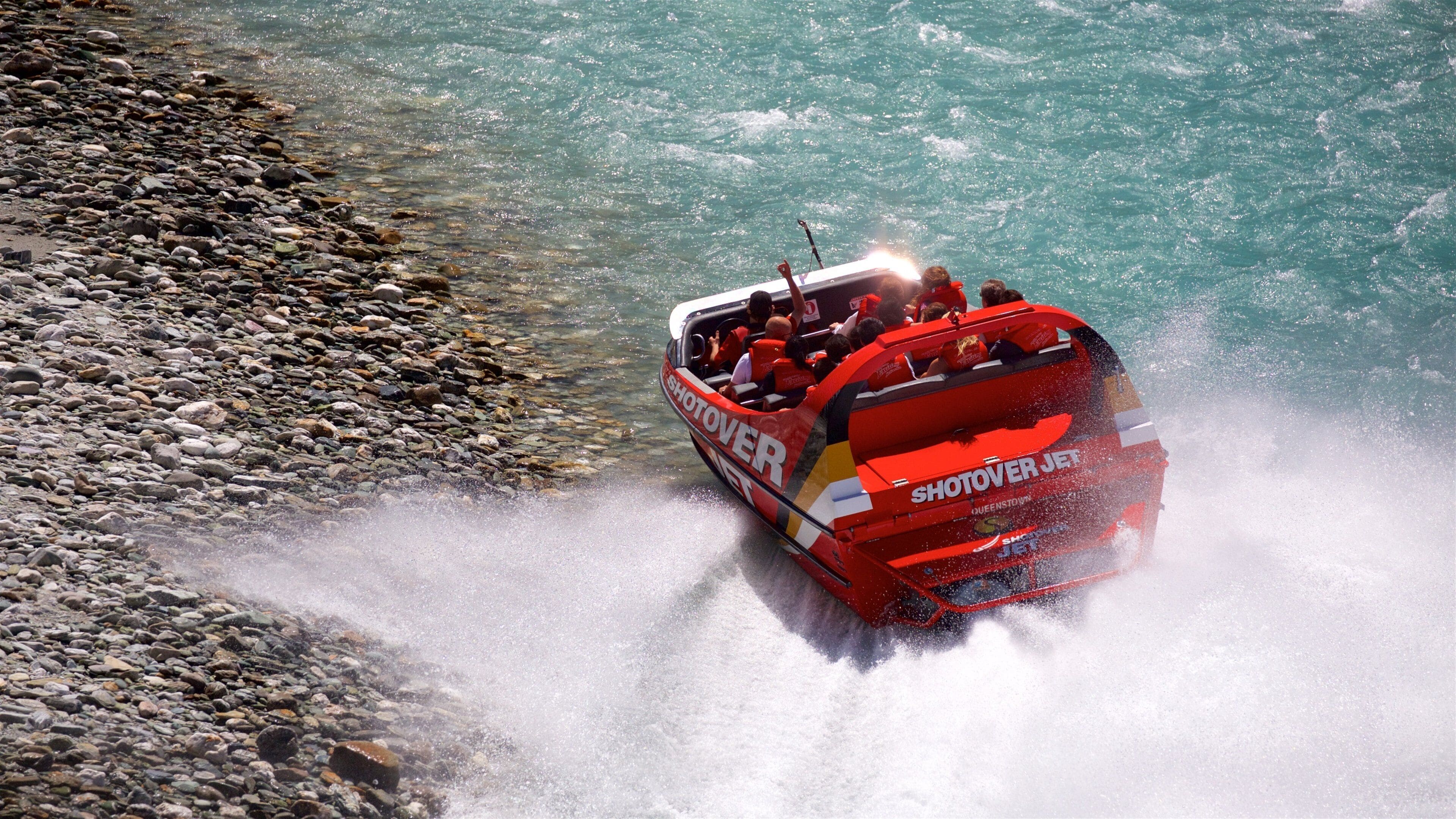 Um barco jacto vermelho chamado "SHOTOVER" movendo-se por águas claras perto de uma margem rochosa.