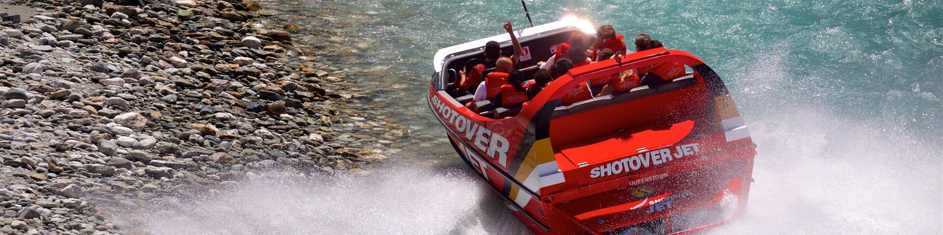 Um barco jacto vermelho chamado "SHOTOVER" movendo-se por águas claras perto de uma margem rochosa.