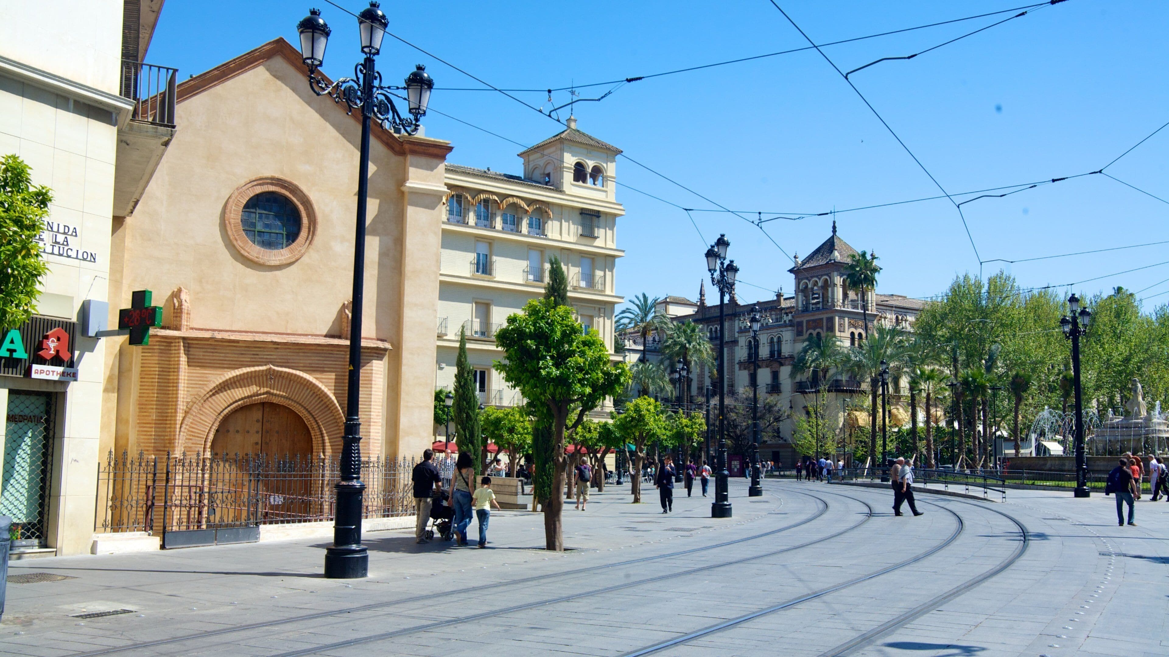 Uma praça urbana ensolarada, com linhas de elétrico, edifícios históricos e pedestres.