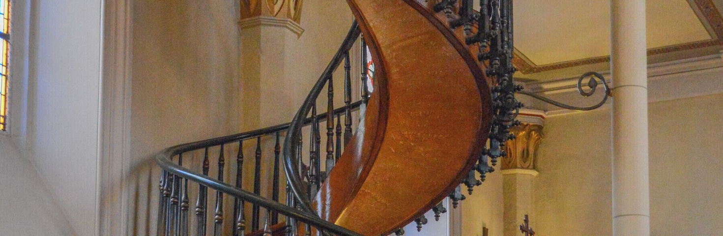 Uma imponente escadaria em espiral com grades ornamentadas e degraus de madeira, dentro de um edifício com janelas de vitrais e estátuas religiosas.
