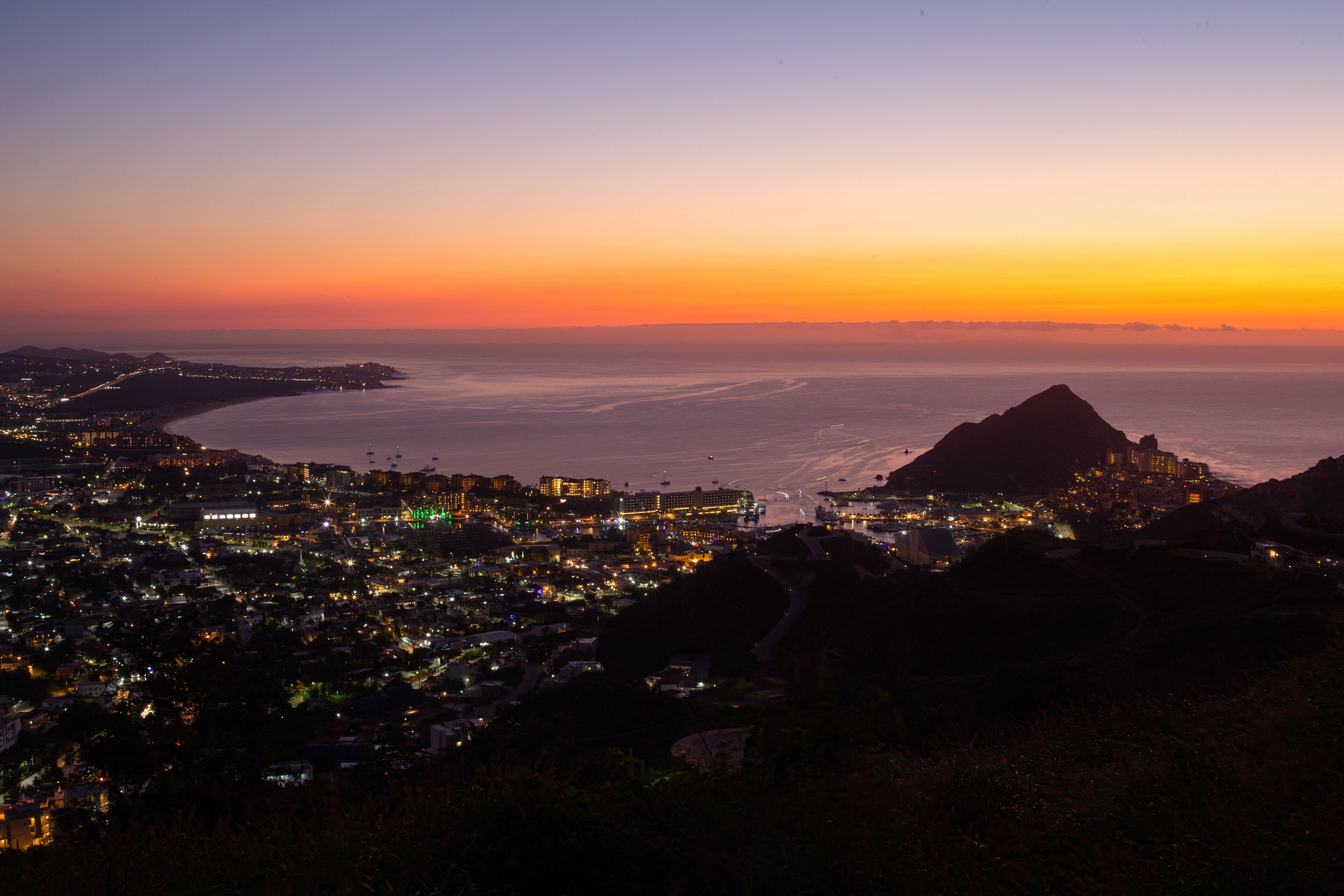 Paisagem costeira urbana ao pôr do sol, com uma montanha imponente e um longo píer que se estende até o oceano.