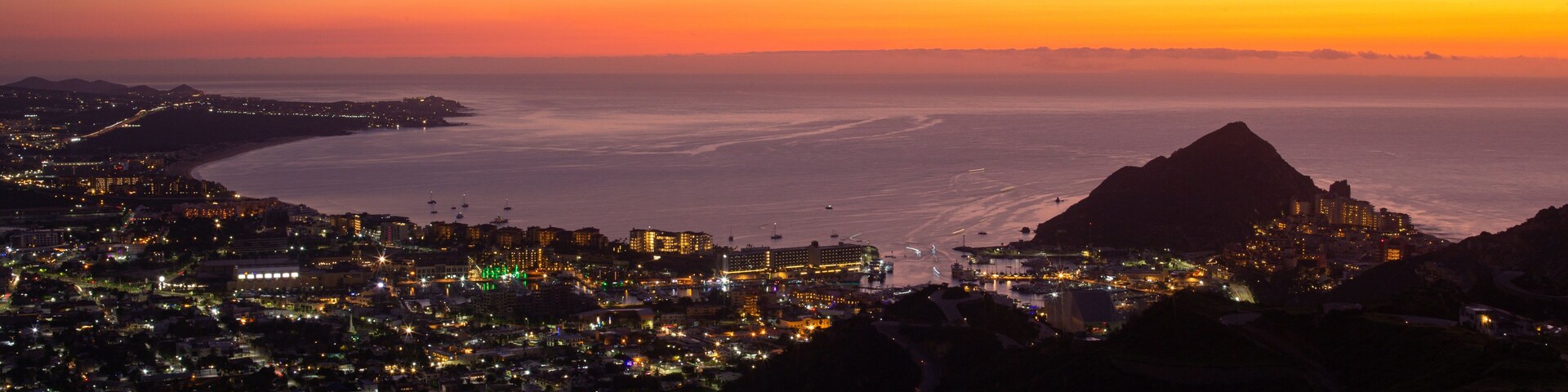 Paisagem costeira urbana ao pôr do sol, com uma montanha imponente e um longo píer que se estende até o oceano.