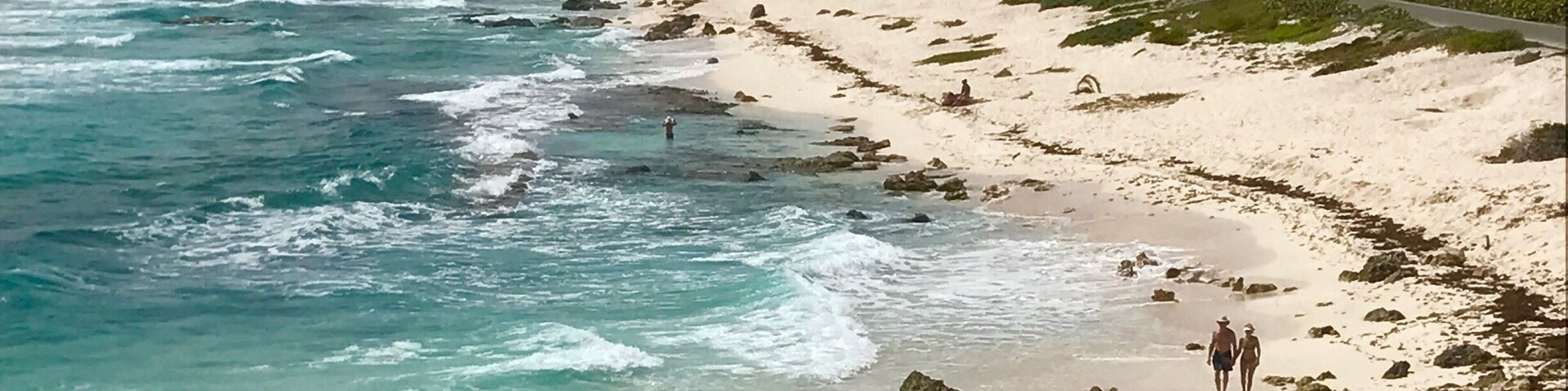 Uma praia com ondas quebrando na costa, algumas pessoas caminhando pela areia e uma estrada que leva a uma aldeia distante.