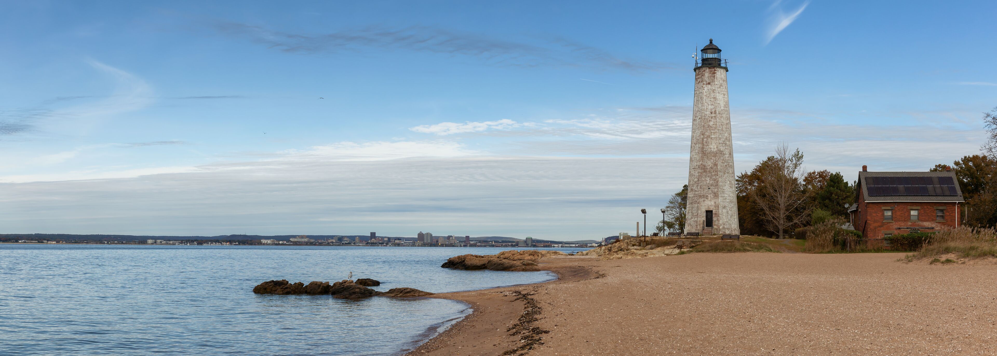 Paisagem costeira com um farol, linha de costa rochosa e um horizonte urbano distante.