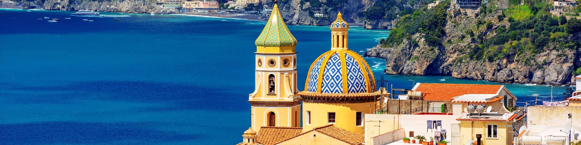 Uma vila costeira com uma igreja destacada, apresentando uma torre verde e uma cúpula azul e branca, rodeada por penhascos íngremes e um mar azul límpido.